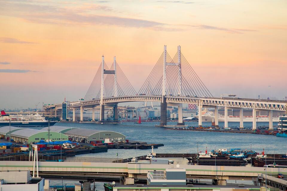 Uma grande ponte suspensa com duas torres, um porto movimentado com navios e contêineres, e um céu ao pôr do sol.