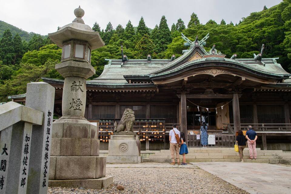 Um templo japonês tradicional com um candeeiro de pedra, uma estátua de leão e visitantes em frente.