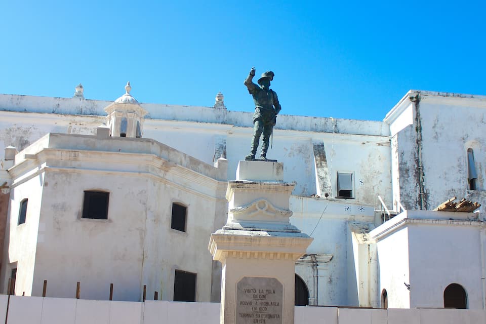 Uma estátua sobre um pedestal em frente a um edifício branco, com um céu azul claro ao fundo.