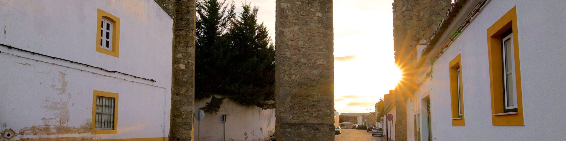 Uma rua de paralelepípedos com arcos de pedra, edifícios brancos com detalhes em amarelo e um pôr do sol ao fundo.