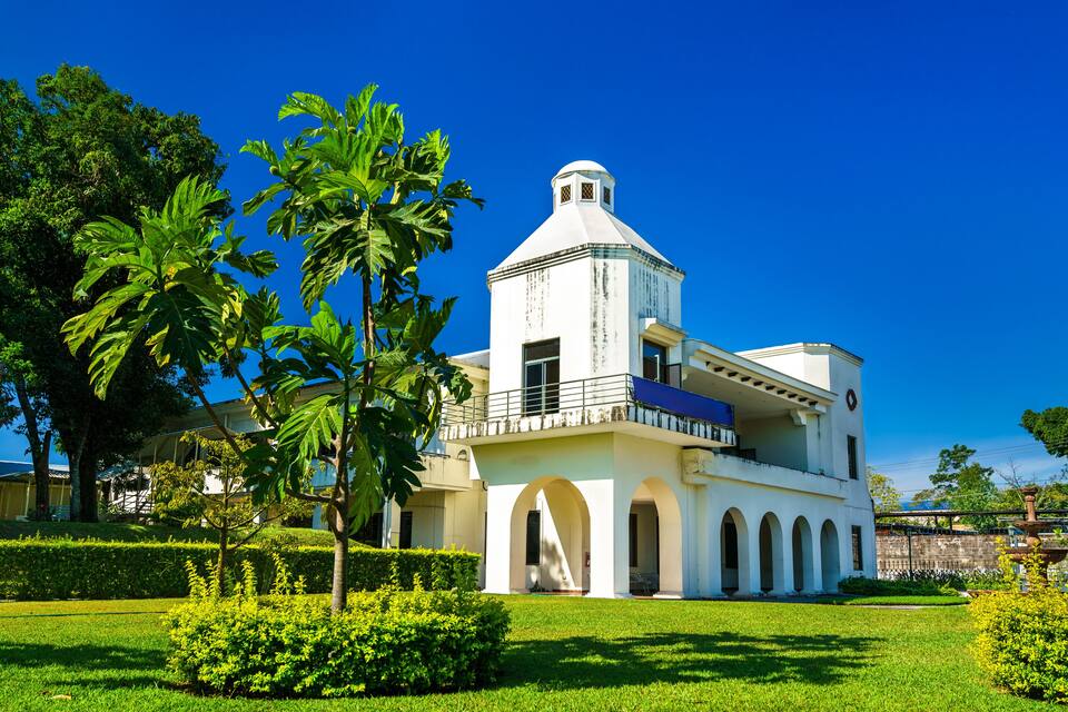 Um edifício de estilo colonial branco com uma cúpula, rodeado por vegetação exuberante e palmeiras.