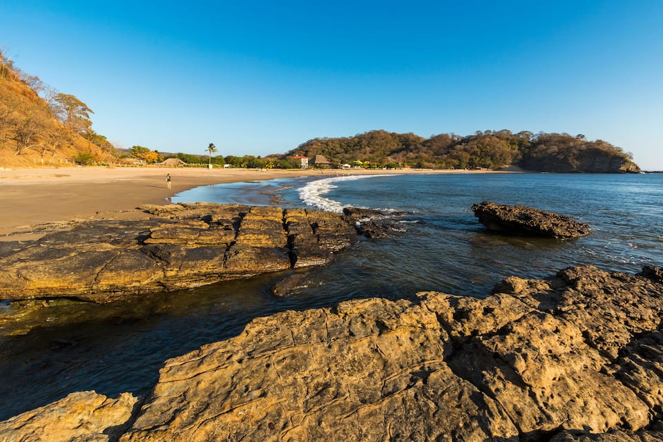 Uma costa rochosa com águas transparentes, uma praia de areia e uma colina distante coberta de árvores.