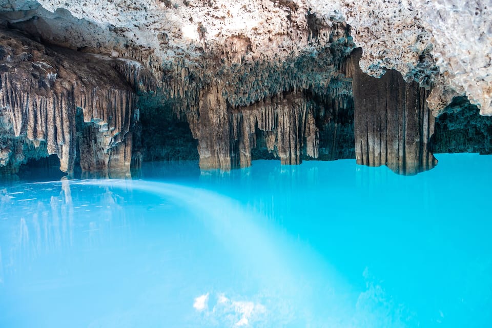 Caverna com estalactites e estalagmites, água azul cristalina e superfície reflexiva.
