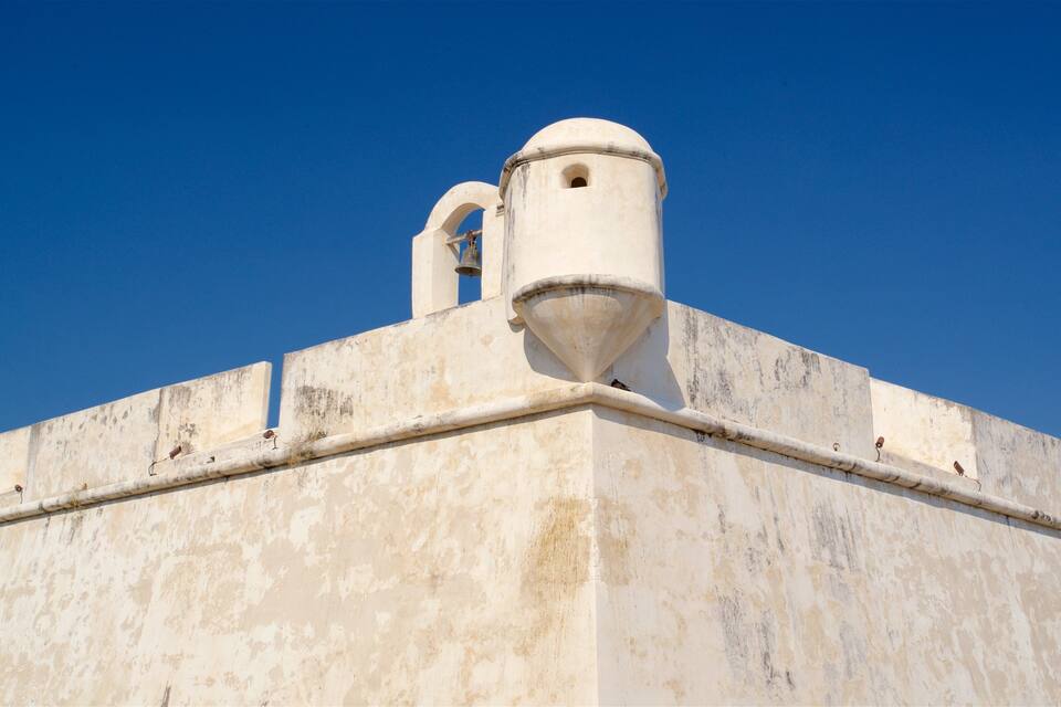 Mural de pedra desgastada com uma estrutura cilíndrica no topo, possivelmente um campanário, contra um céu azul límpido.