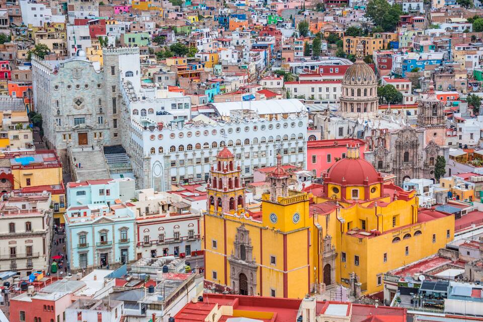 Uma área urbana densamente povoada com edifícios coloridos, incluindo uma igreja amarela imponente com cúpula vermelha e um edifício branco com torre de relógio.
