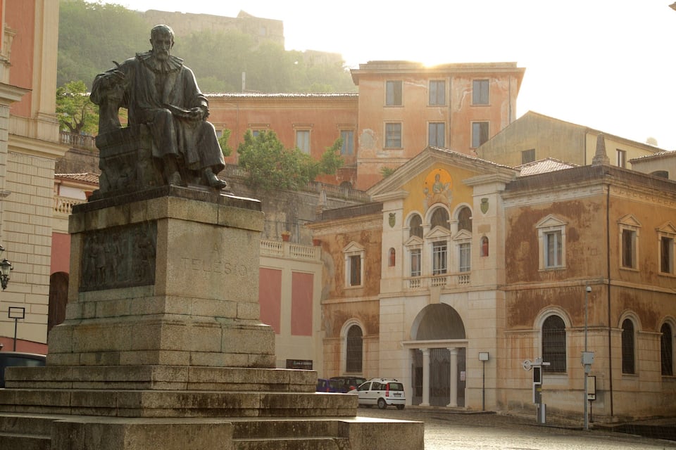 Estátua de um homem sobre um pedestal numa praça, com edifícios históricos ao fundo.