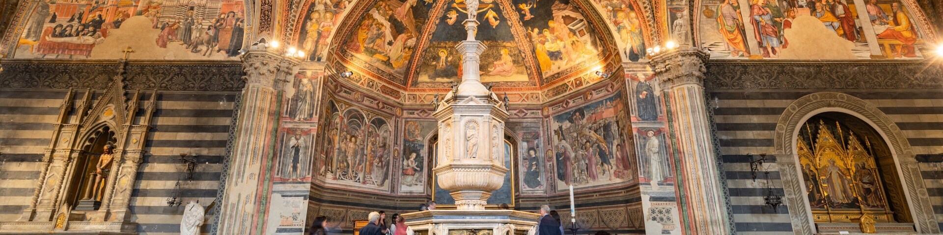 Interior imponente de uma catedral, com afrescos detalhados, um altar central ricamente decorado e visitantes apreciando as obras de arte.