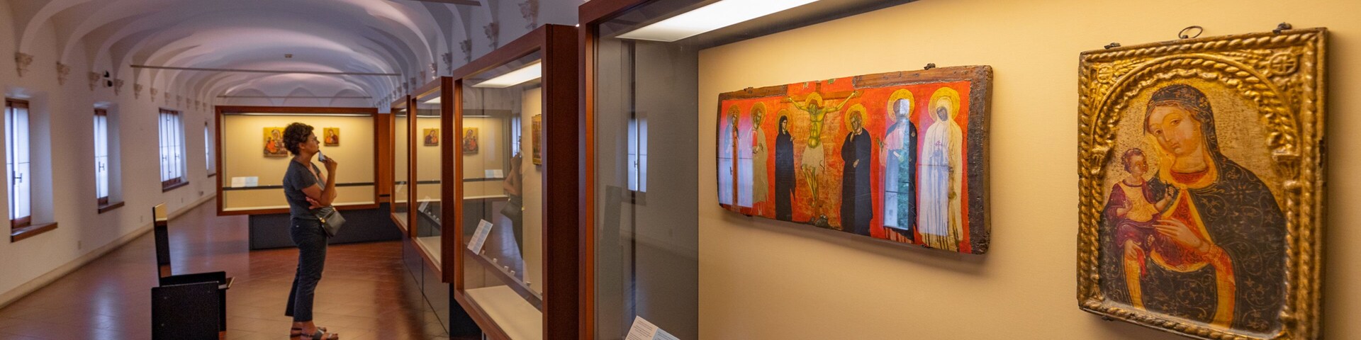 Um corredor de museu com obras de arte expostas, incluindo uma imagem religiosa e um tríptico.