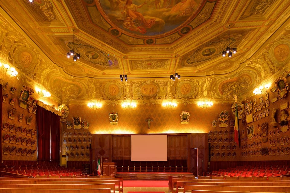 Um teatro ornamentado com teto dourado, cortinas vermelhas e fileiras de assentos.