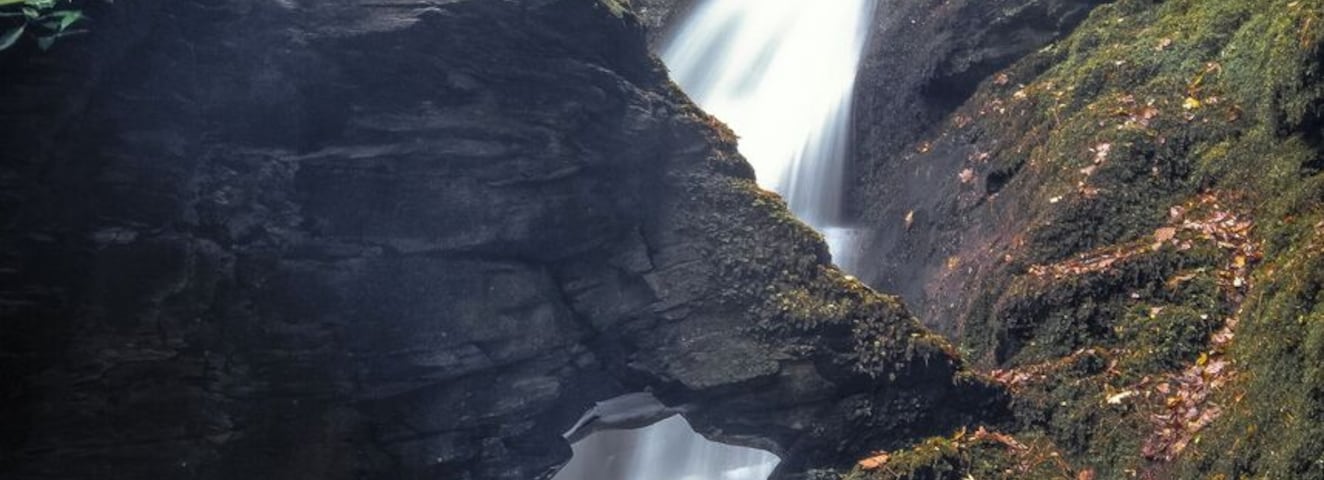 Uma cascata desce por um desfiladeiro rochoso com paredes cobertas de musgo e uma pequena piscina na base.