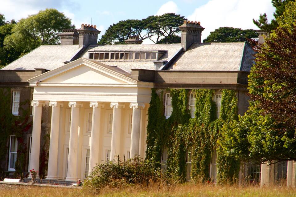 Um edifício clássico com pórtico e colunas, rodeado por vegetação e árvores.