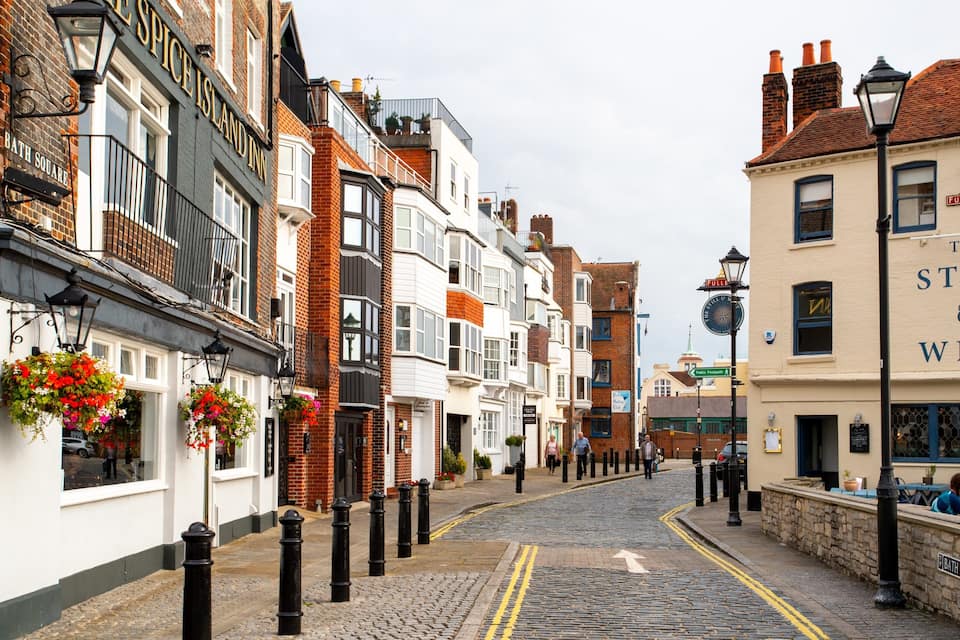 Uma rua de calçada adornada por edifícios históricos, incluindo um pub chamado 'The Still & West' e 'Bath Square'.