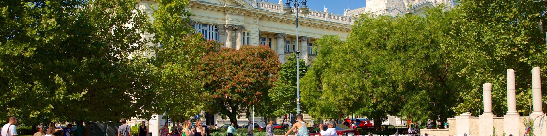 Uma praça pública com uma fonte, árvores e um edifício grande e ornamentado ao fundo.