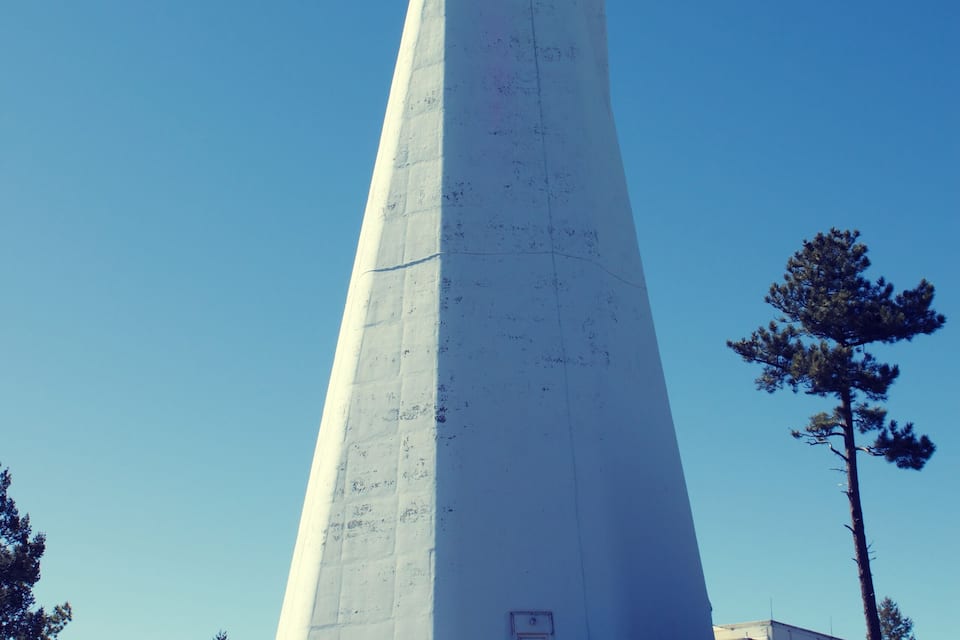 Uma torre branca e alta com topo pontiagudo, cercada por vegetação rasteira e um céu azul límpido.