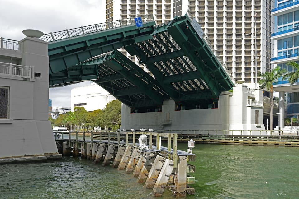 Uma ponte para pedestres verde sobre a água, com um edifício branco ao fundo.
