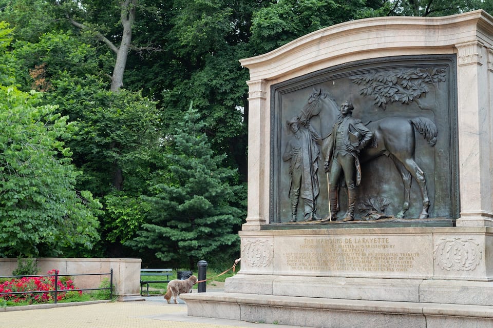 Estátua equestre em bronze de uma figura histórica, rodeada por vegetação exuberante e um banco de parque.