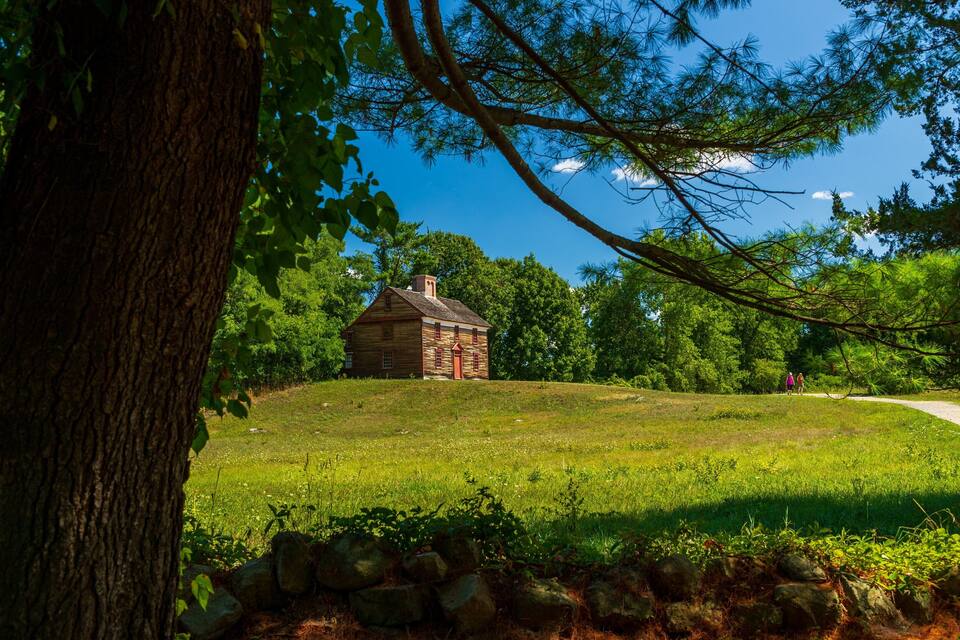 Uma cabana de toras histórica em um campo gramado, com um muro de pedra em primeiro plano e um céu azul límpido.