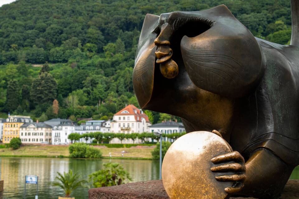 Escultura em bronze de um elefante grande segurando um globo, com um rio e edifícios ao fundo.