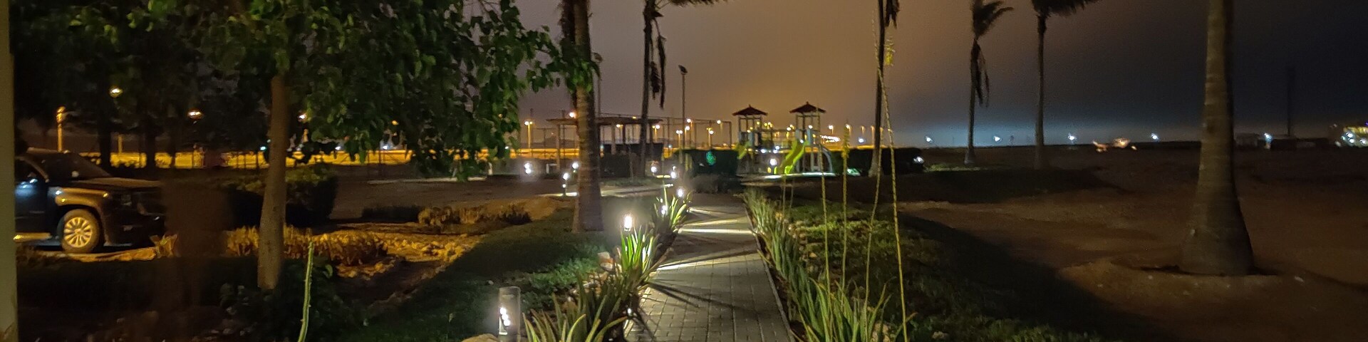 Uma passagem noturna bordeada por palmeiras e plantas de aloe, levando a uma praia com luzes ao longe.