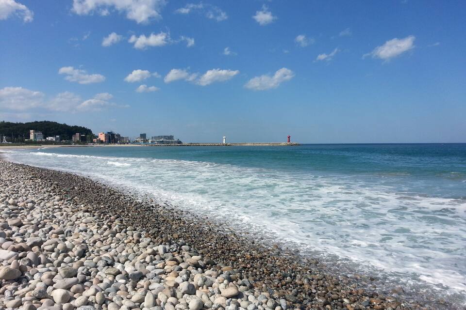 Uma praia de pedras com ondas, um farol e edifícios ao fundo.
