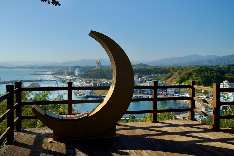 Um deck de madeira com uma escultura em formato de crescente, com vista para um horizonte costeiro e montanhas ao fundo.