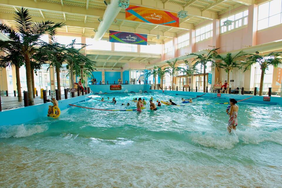 Parque aquático interno com várias piscinas, palmeiras e pessoas nadando e brincando.