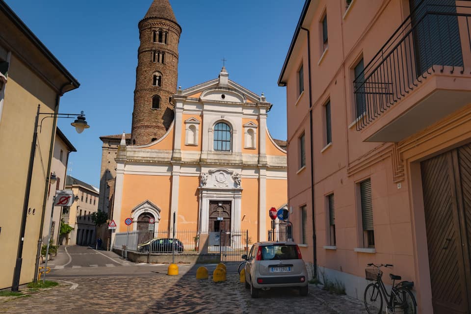 Uma rua de paralelepípedos com uma igreja, destacando-se uma torre alta de sino e um carro estacionado.