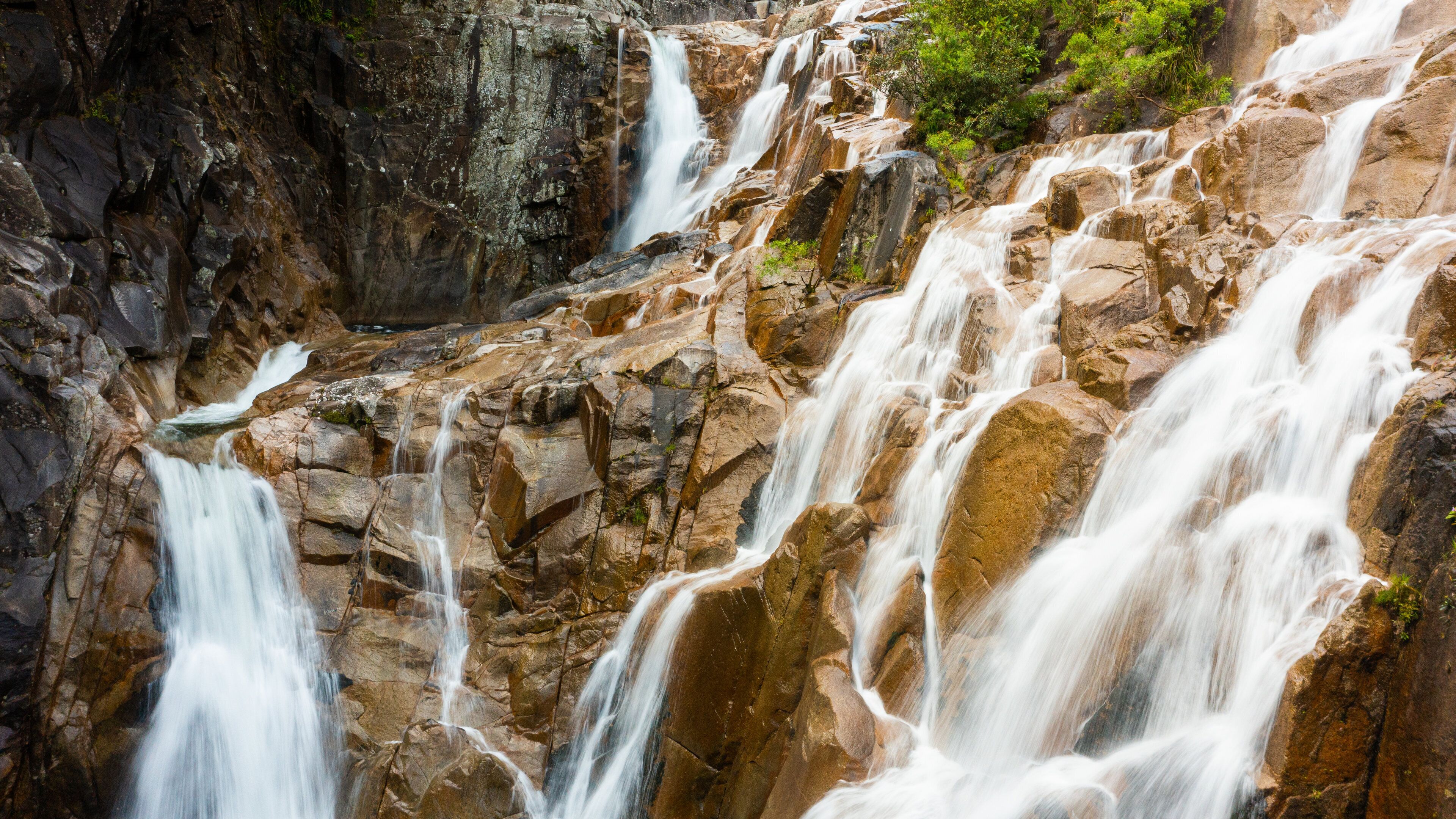 Uma cascata multifacetada desce sobre terreno rochoso, com vegetação exuberante no topo.