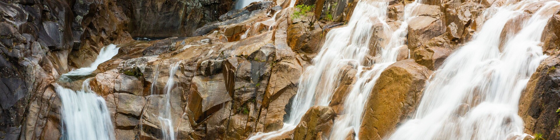 Uma cascata multifacetada desce sobre terreno rochoso, com vegetação exuberante no topo.