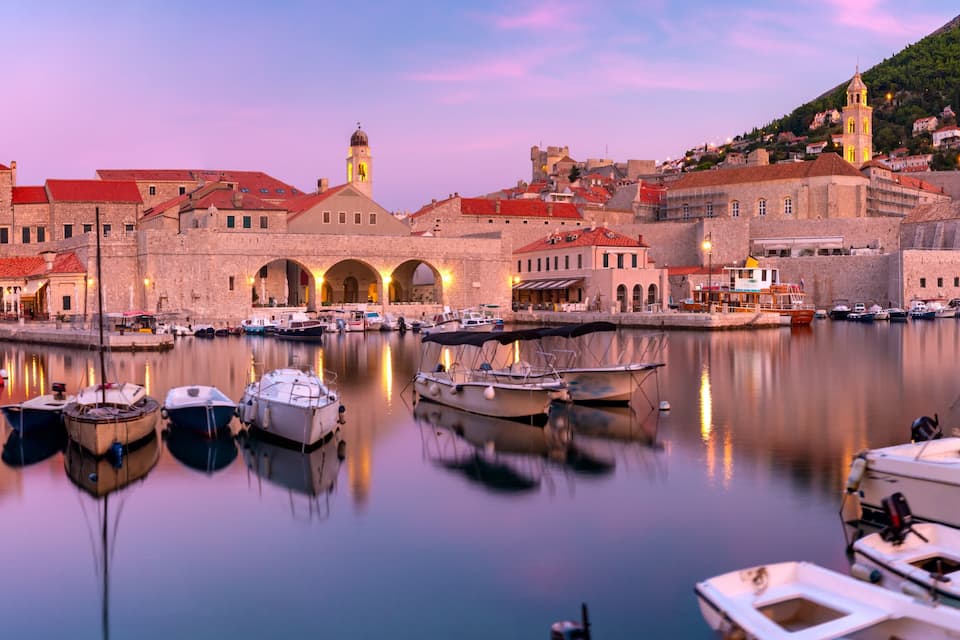 Um porto histórico com barcos, um edifício imponente com torre do relógio e um morro com casas.