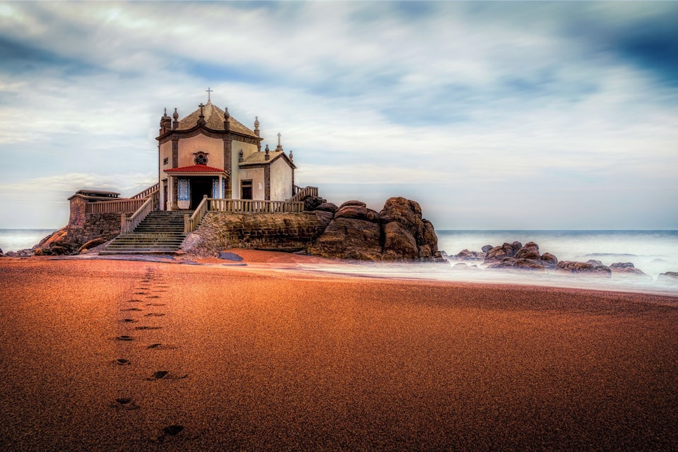 Uma igreja costeira com telhado de telhas vermelhas, situada em um afloramento rochoso com uma praia arenosa em primeiro plano.