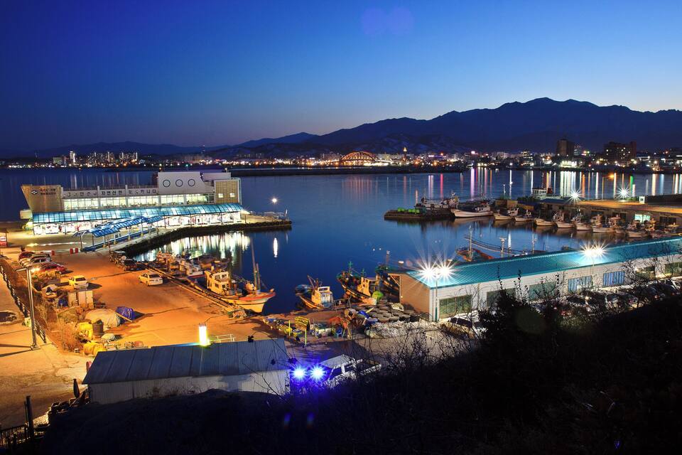 Marina à beira-mar ao entardecer, com barcos atracados, uma ponte ao fundo e o horizonte urbano refletido na água.