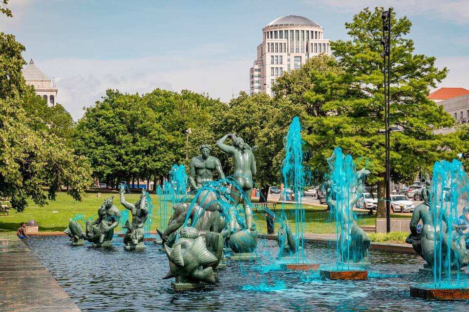 Um parque público com uma fonte adornada por esculturas de bronze de figuras mitológicas, rodeado por árvores e um grande edifício ao fundo.