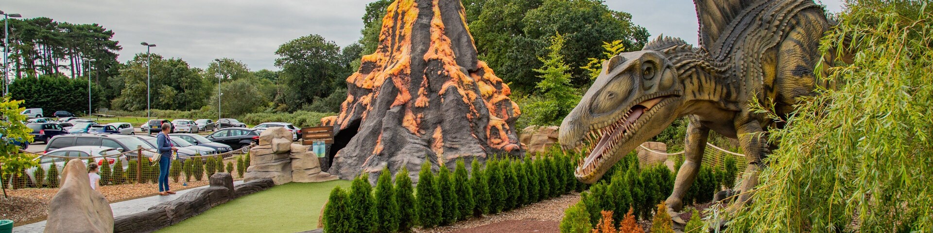 Um parque com tema de dinossauros, apresentando uma grande estátua de dinossauro, um vulcão e um estacionamento.