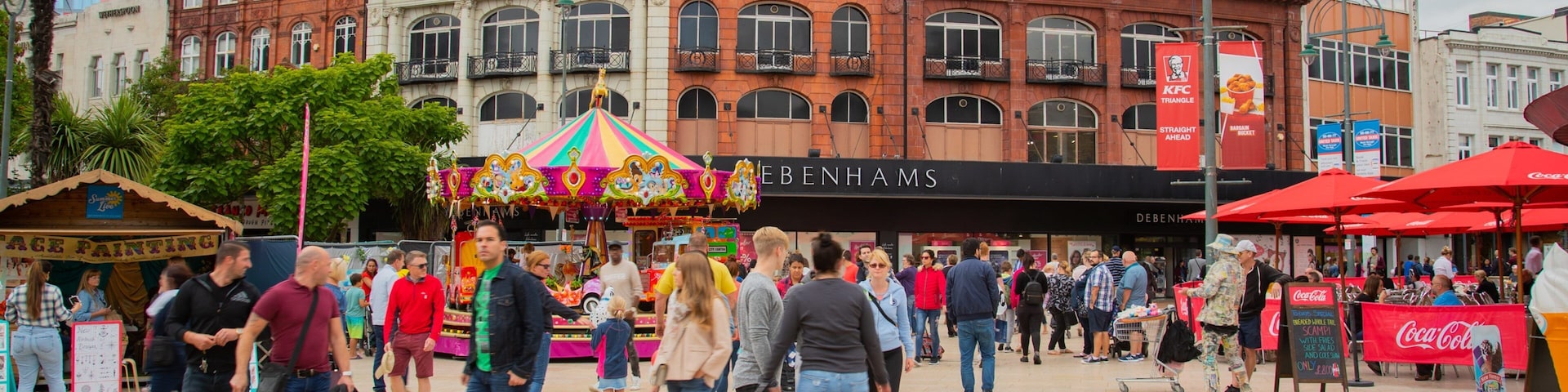 Uma praça pública movimentada com um carrossel, pessoas caminhando e edifícios com placas como 'DEBENHAMS' e 'KFC'.