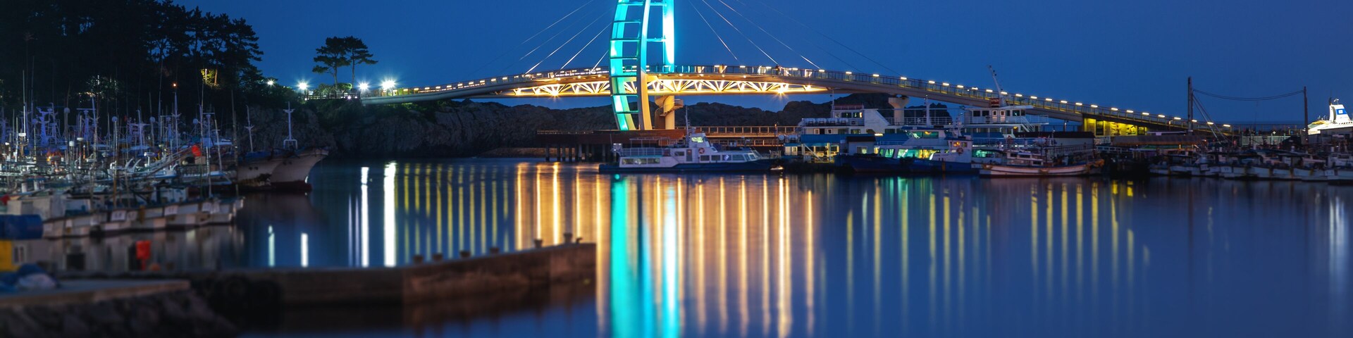 Uma ponte com um design azul e branco característico, refletida em águas calmas, rodeada por barcos e um porto de recreio.