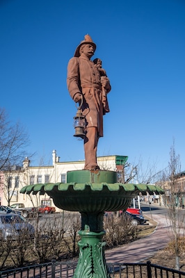 Estátua de um homem com chapéu segurando um candeeiro, sobre um pedestal verde.