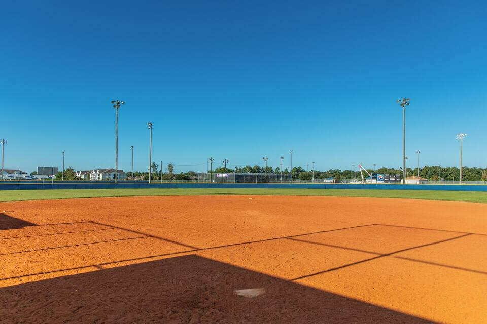 Um campo de beisebol com céu azul, refletores e placar ao fundo.