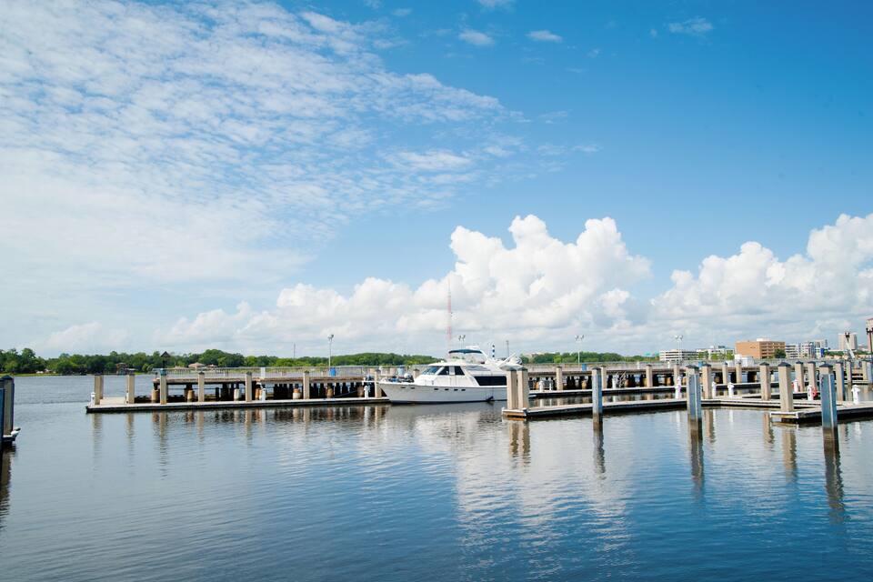 Uma marina com um iate branco atracado em um cais, rodeada por águas calmas e um céu claro com algumas nuvens.