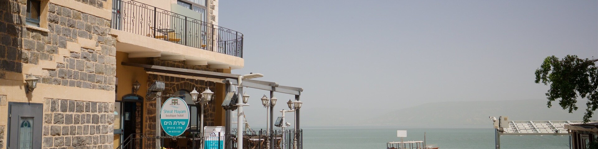 Um edifício de pedra com varanda, placa indicando o hotel boutique Shirat Hayam e vista do mar com um barco ao longe.