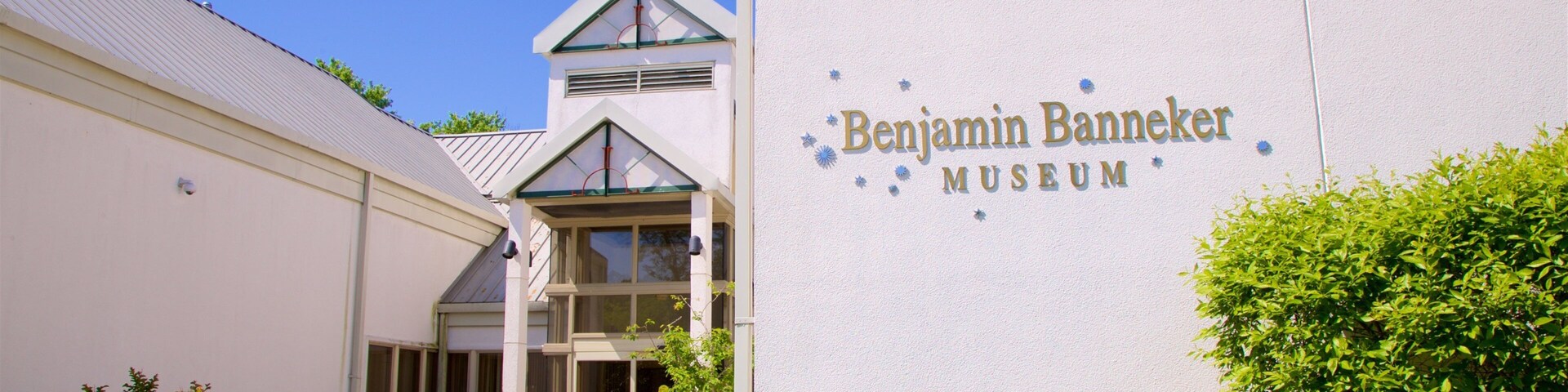 Exterior do Museu Benjamin Banneker com céu claro, um edifício branco e uma pequena área de jardim com bancos.