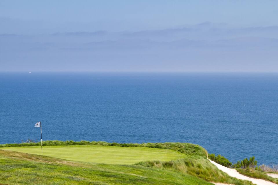 Um campo de golfe com uma bandeira, um green e vista para o oceano.