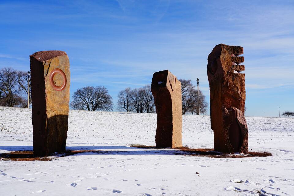 Três esculturas de metal enferrujado em um campo nevado, com árvores secas ao fundo.