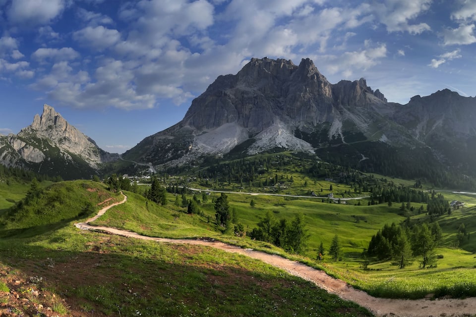 Paisagem montanhosa com um caminho sinuoso, campos verdes e um pico imponente.