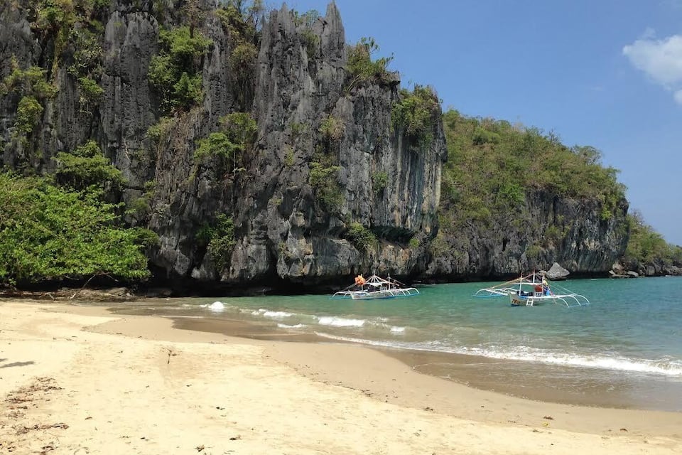 Uma praia com águas turquesa cristalinas, falésias rochosas e barcos de pesca tradicionais.