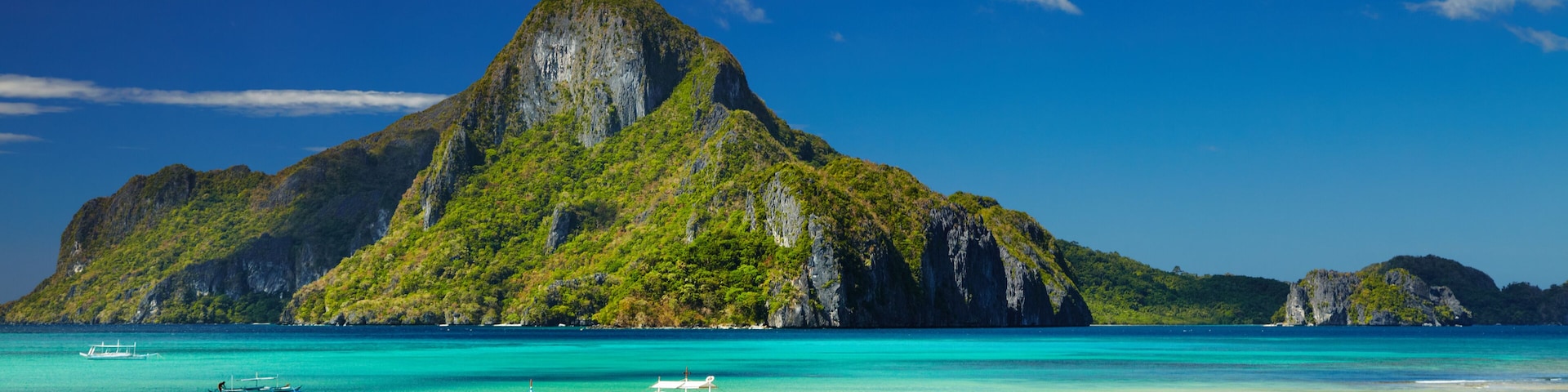 Uma praia tropical com águas turquesa cristalinas, pequenos barcos e um imponente pico de calcário.