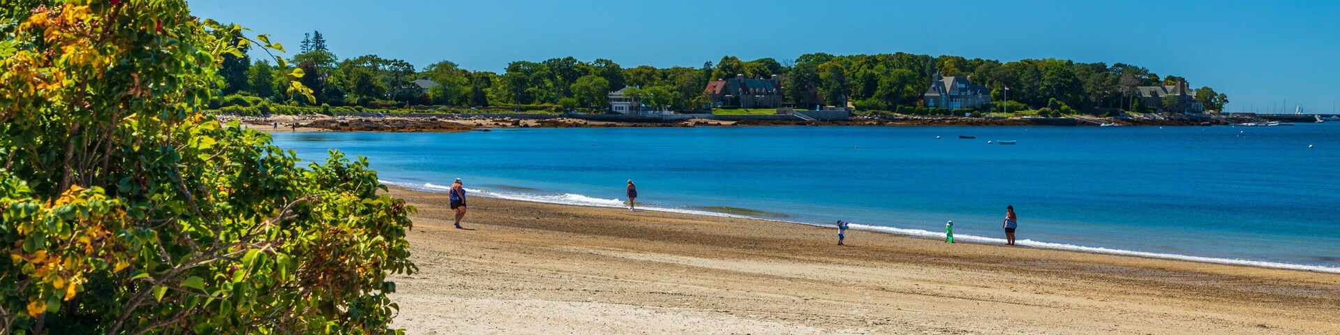 Uma praia arenosa com céu azul límpido, mar calmo e casas ao fundo.