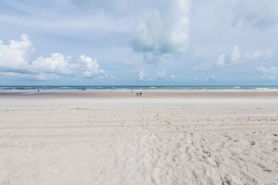Uma extensa praia de areia com ondas suaves, algumas pessoas à distância e céu parcialmente nublado.