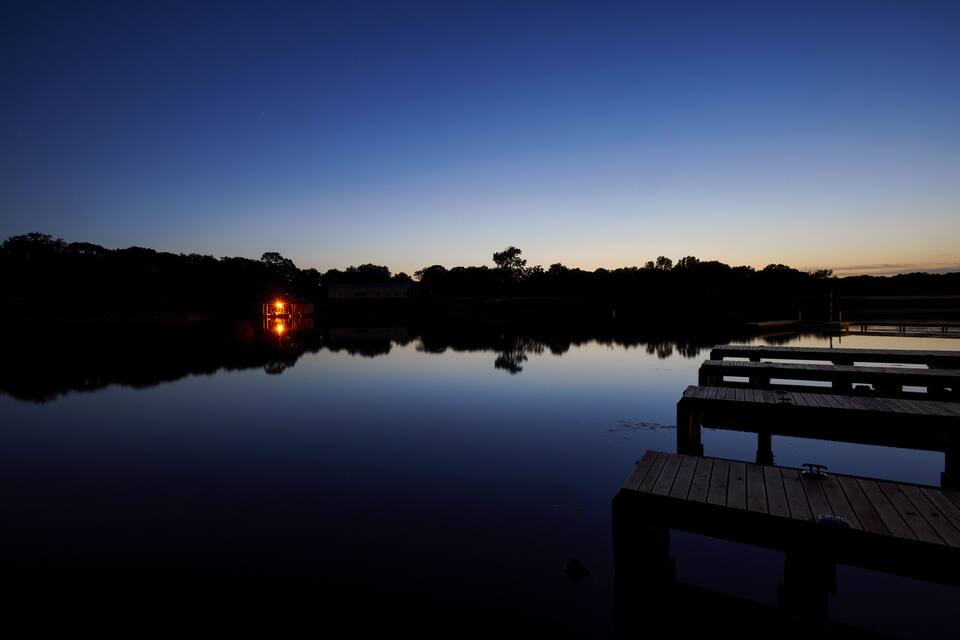 Um lago calmo ao entardecer, com um píer em primeiro plano e um barco iluminado à distância.