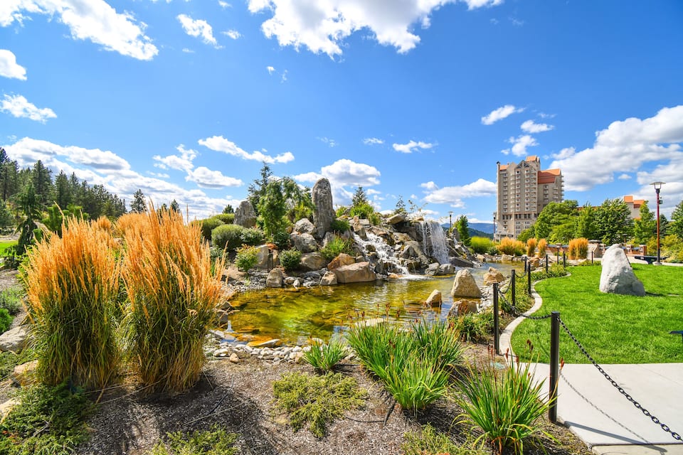 Um jardim com paisagismo, um lago, pedras e um edifício ao fundo.